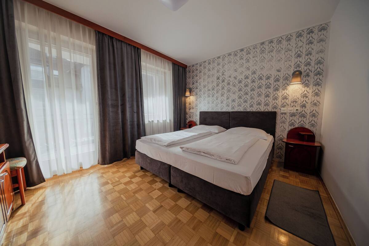 Quarto com cama, cortinas, mesa de cabeceira, luminária na parede e papel de parede estampado.