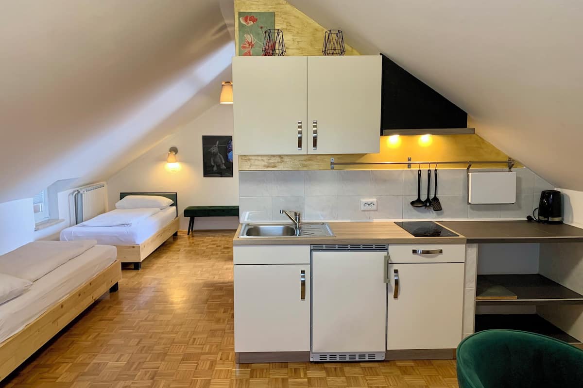 Quarto compacto e bem iluminado com cozinha compacta, duas camas, um banco e uma cadeira verde.