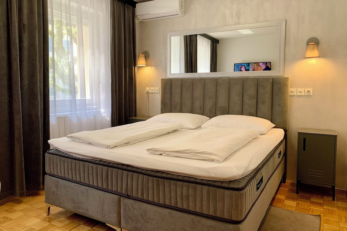 Cama bem arrumada em quarto de hotel, com cabeceira, ar condicionado, mesa de cabeceira, espelho e um pequeno armário.