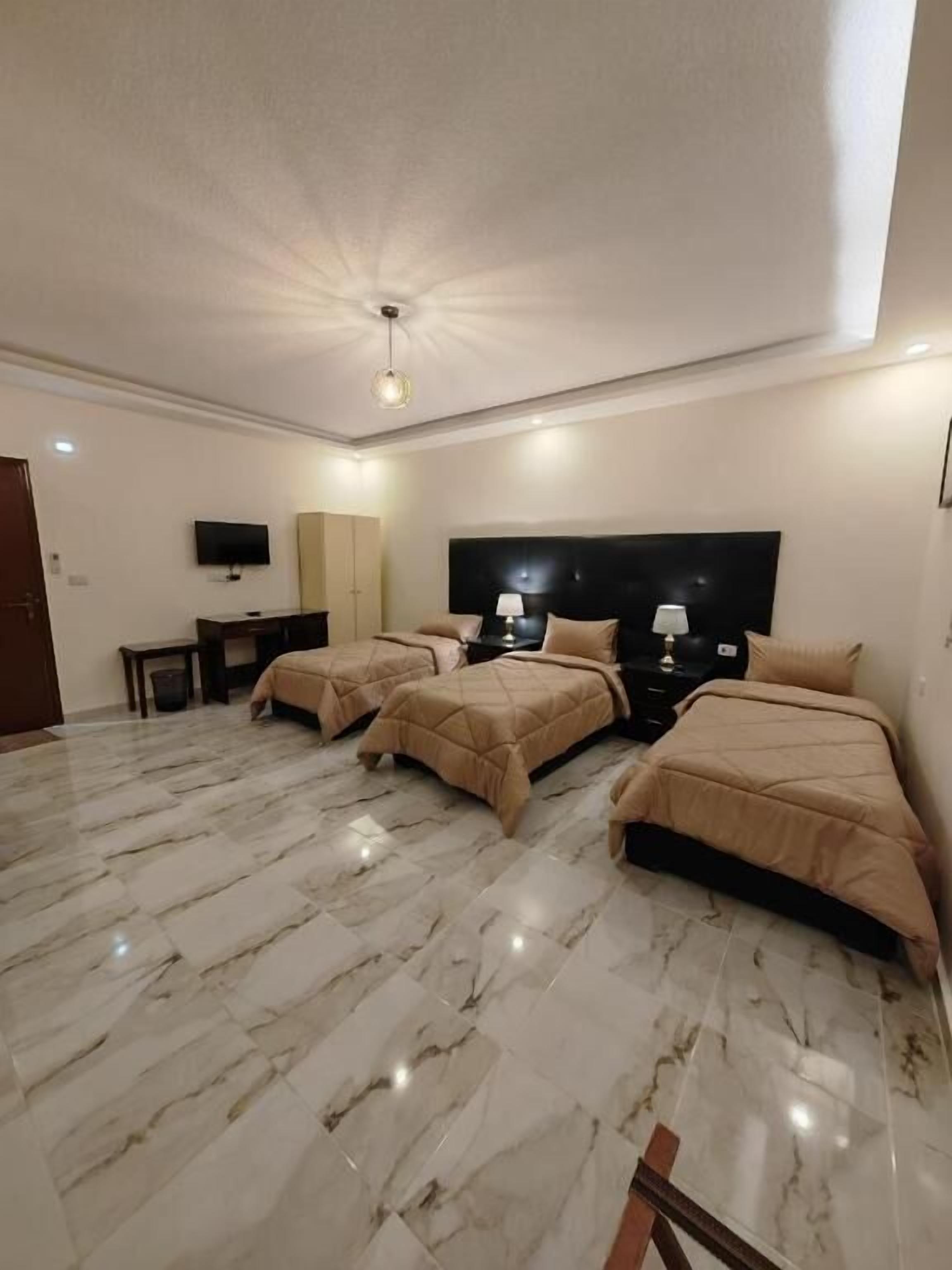 Quarto de hotel com uma cama grande, uma cama menor, uma escrivaninha, uma cadeira, uma televisão e uma luminária no teto.