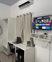 Quarto moderno com escrivaninha branca, um laptop, televisão na parede e uma pequena planta.