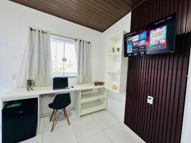 Quarto com escrivaninha branca, cadeira preta, televisão fixada em um painel de madeira e janela com cortinas.