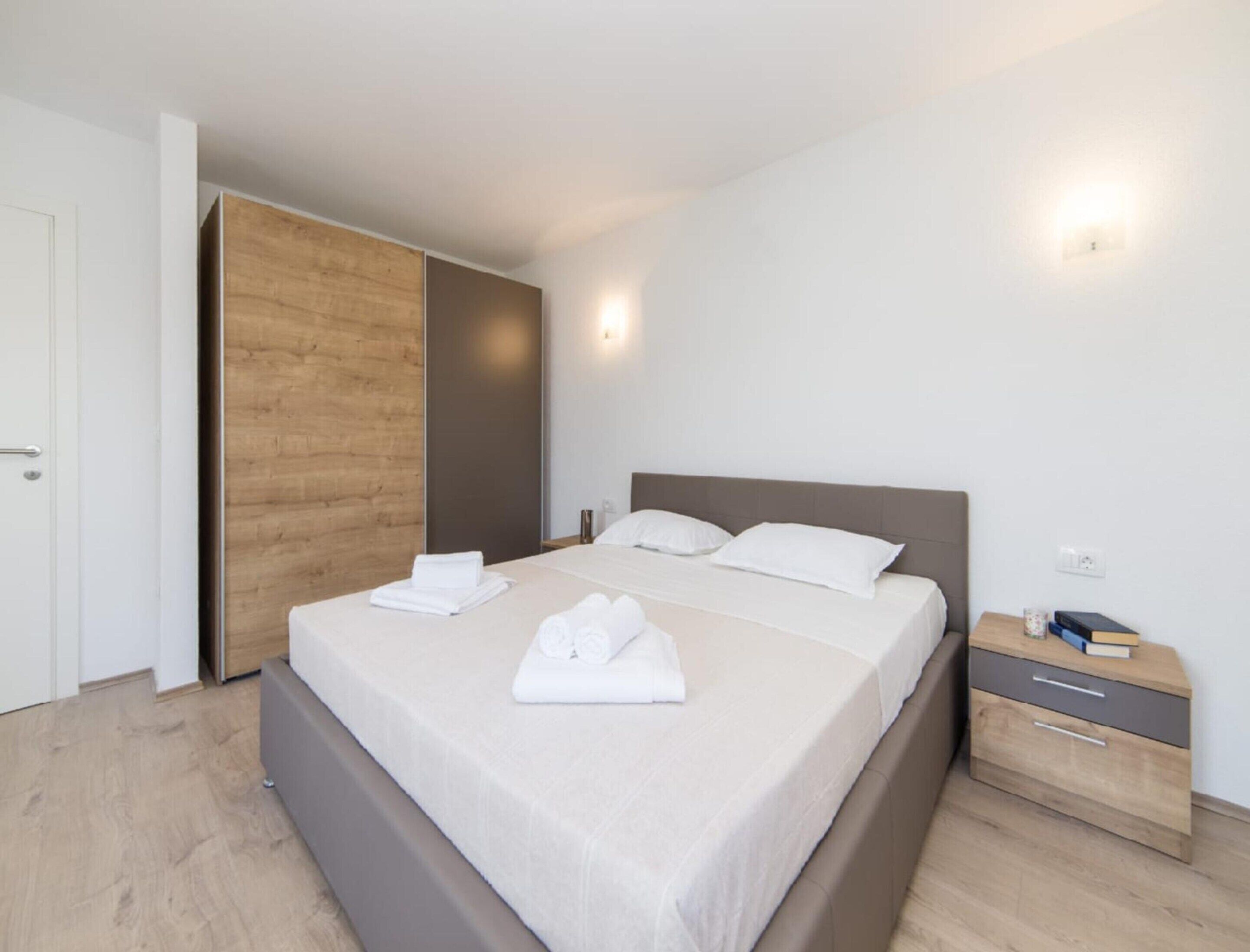 Quarto de hotel moderno com uma cama grande, um guarda-roupa de madeira, mesa de cabeceira com abajur, e criado-mudo com um livro e um telefone.