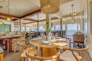 Uma sala de jantar espaçosa, com pé direito alto, janelas amplas e vista para o mar.