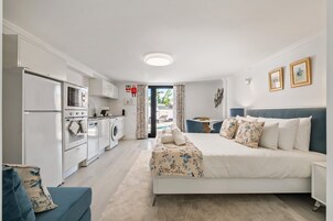 Quarto de hotel moderno com cozinha compacta, cama com roupa de cama floral, sofá azul e vista para o exterior.