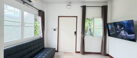 Quarto de hotel com sofá de couro preto, televisão de tela plana na parede, porta branca e janela com cortinas.