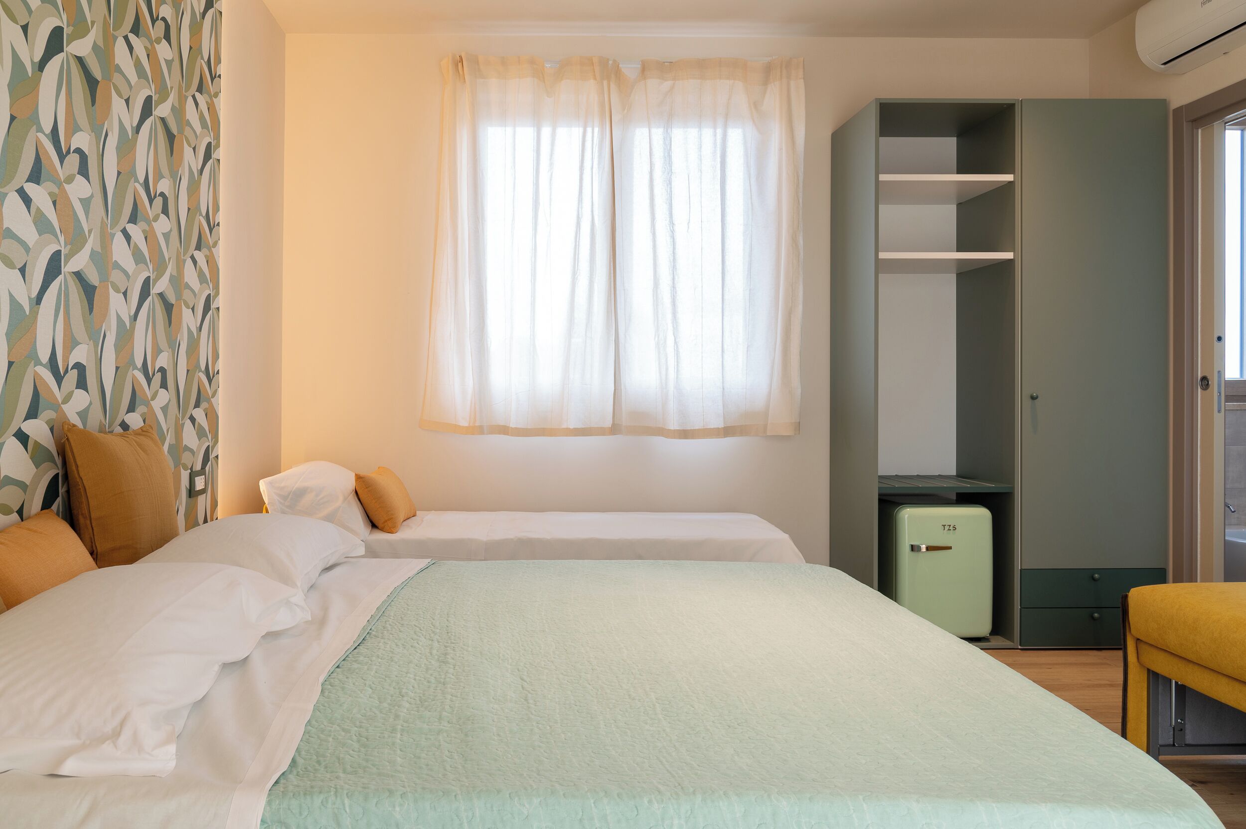 Quarto com duas camas, um guarda-roupa, um eletrodoméstico pequeno verde e uma janela com cortinas leves.