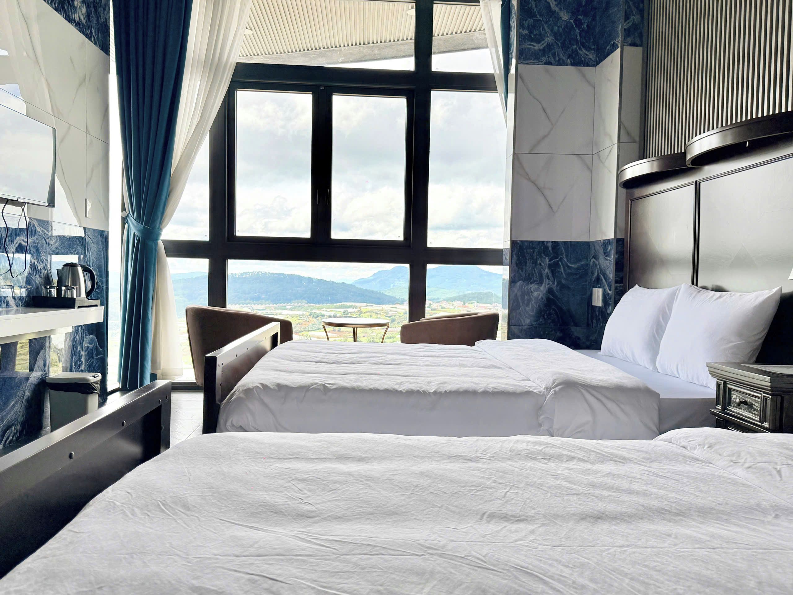 Quarto de hotel com duas camas, uma mesa de jantar e vista para montanhas por grandes janelas.