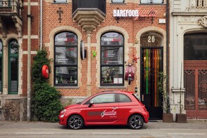 Um carro vermelho estacionado em frente a um hotel com fachada de tijolos, janelas arqueadas grandes e um letreiro de neon.