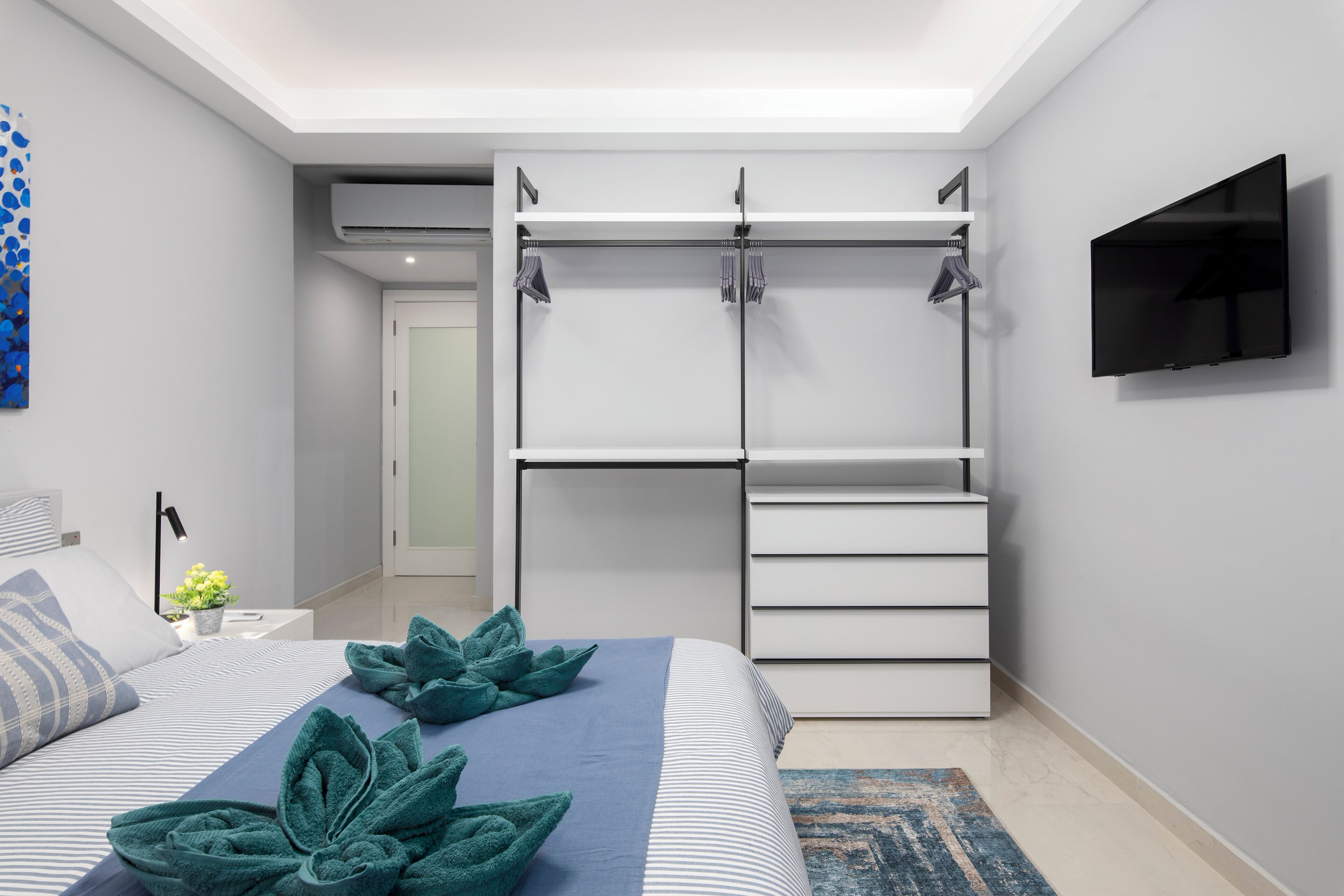 Quarto moderno com cama, roupeiro embutido, televisão de tela plana e pequena mesa com abajur e flores.