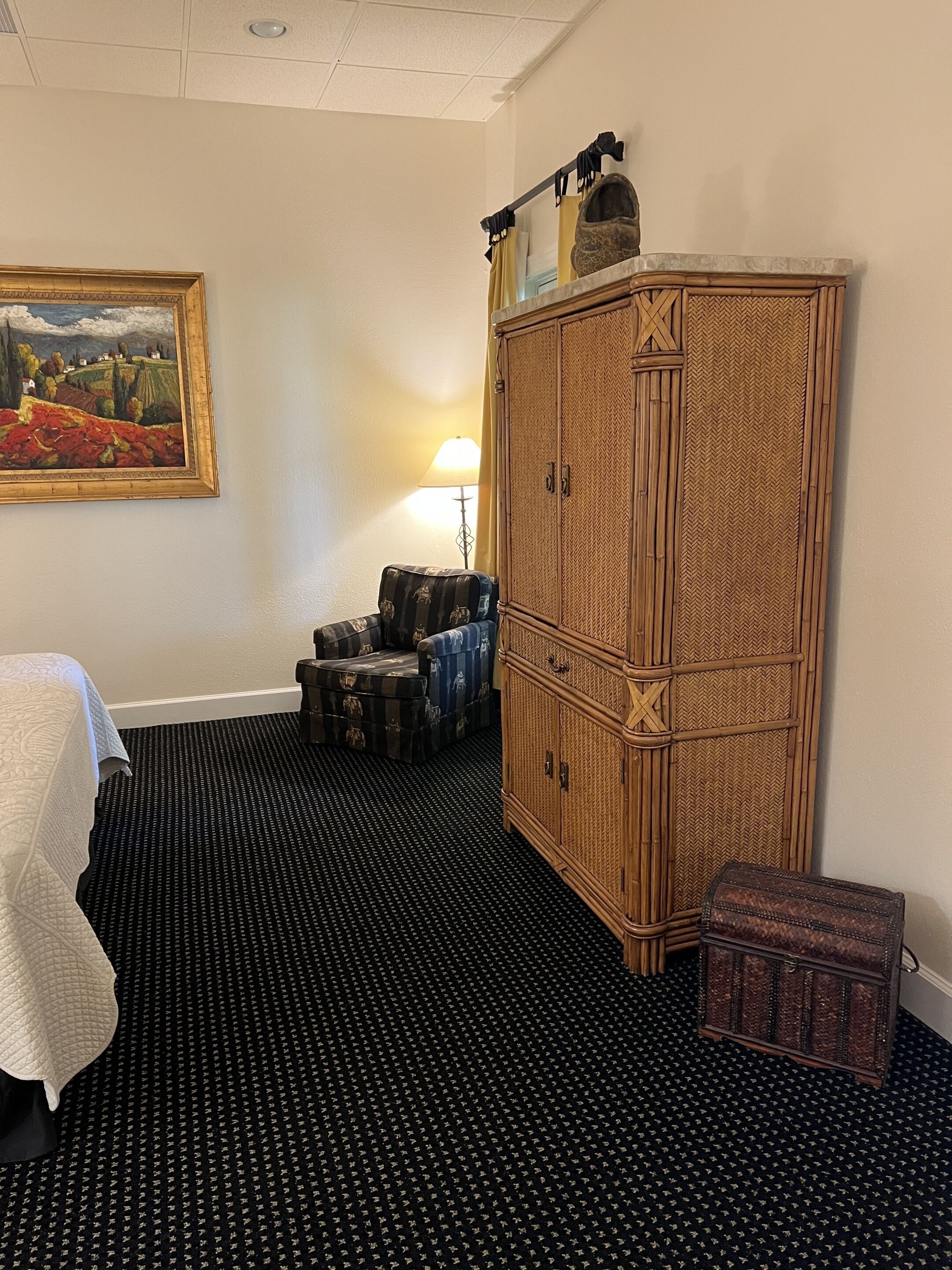 Quarto de hotel com cama, um armário grande de fibra natural, uma mesinha com abajur e um quadro na parede.