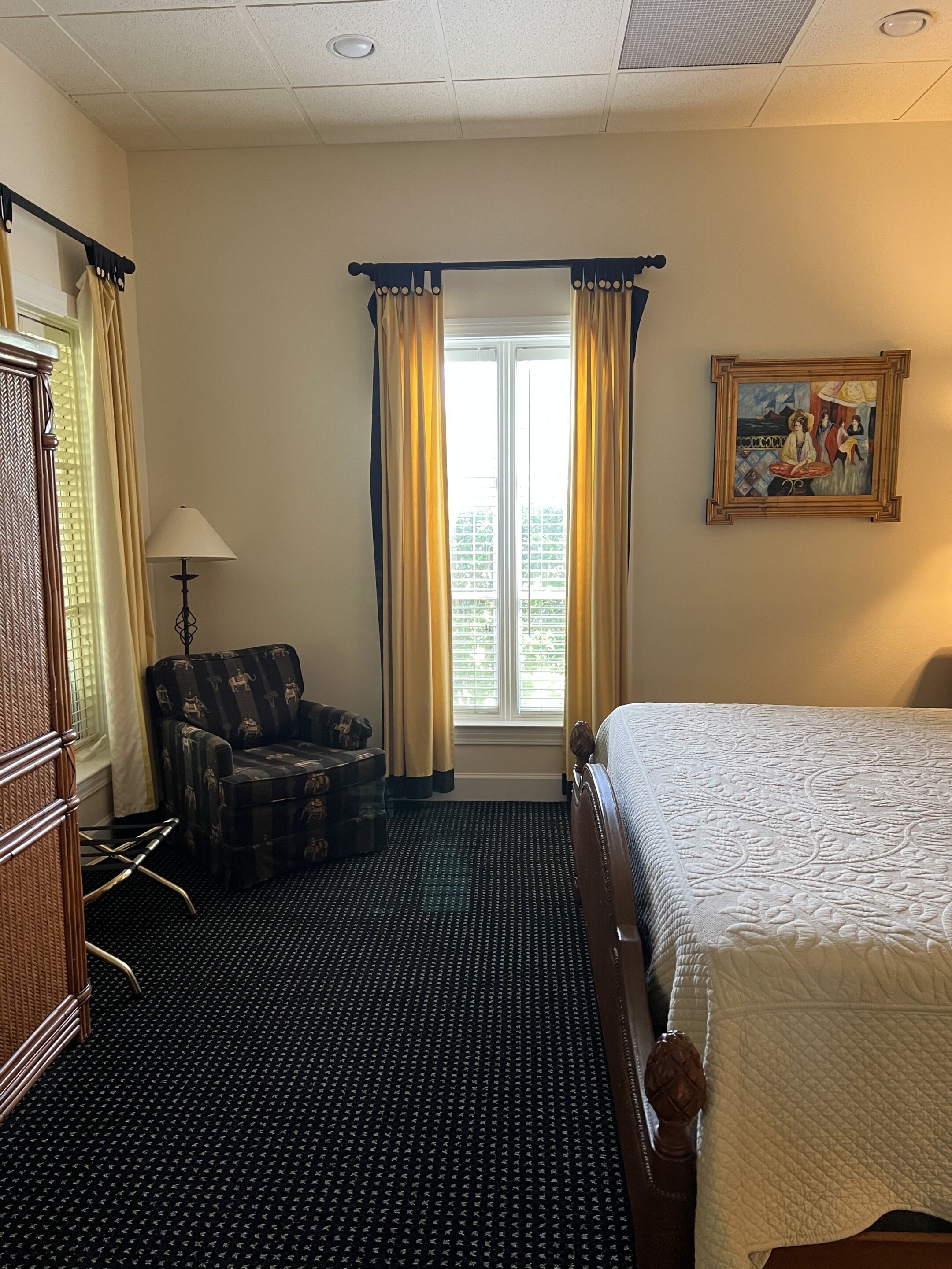 Quarto de hotel com cama, cadeira, escrivaninha e janela com cortinas.