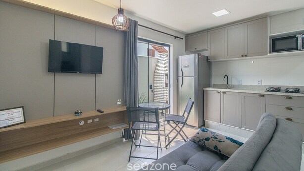 Uma sala de estar moderna com um sofá, uma mesa de jantar com cadeiras, uma televisão de tela plana na parede e uma área de cozinha com armários e micro-ondas.