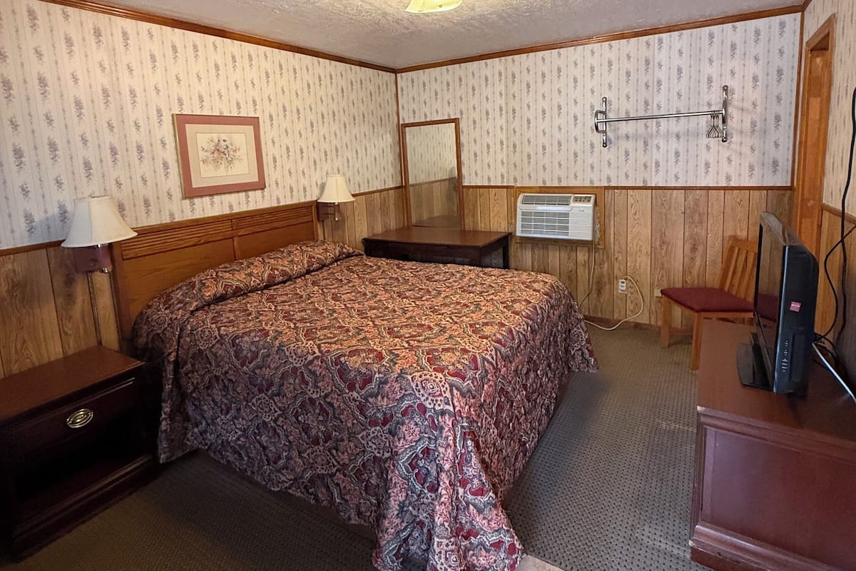 Um quarto com uma cama, uma escrivaninha, uma cadeira, uma televisão e um espelho.