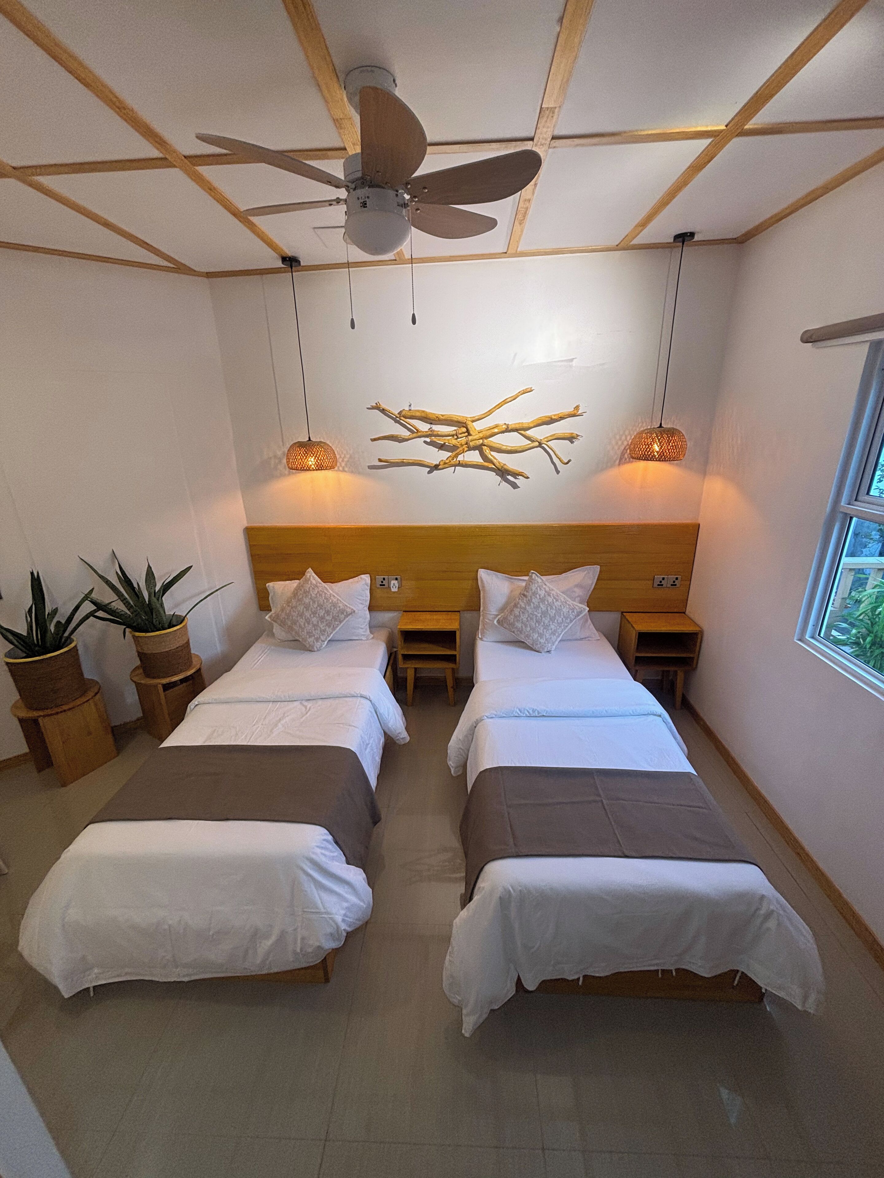 Quarto de hotel com duas camas, um ventilador de teto, cabeceira de madeira, mesas de cabeceira e duas luminárias pendentes.
