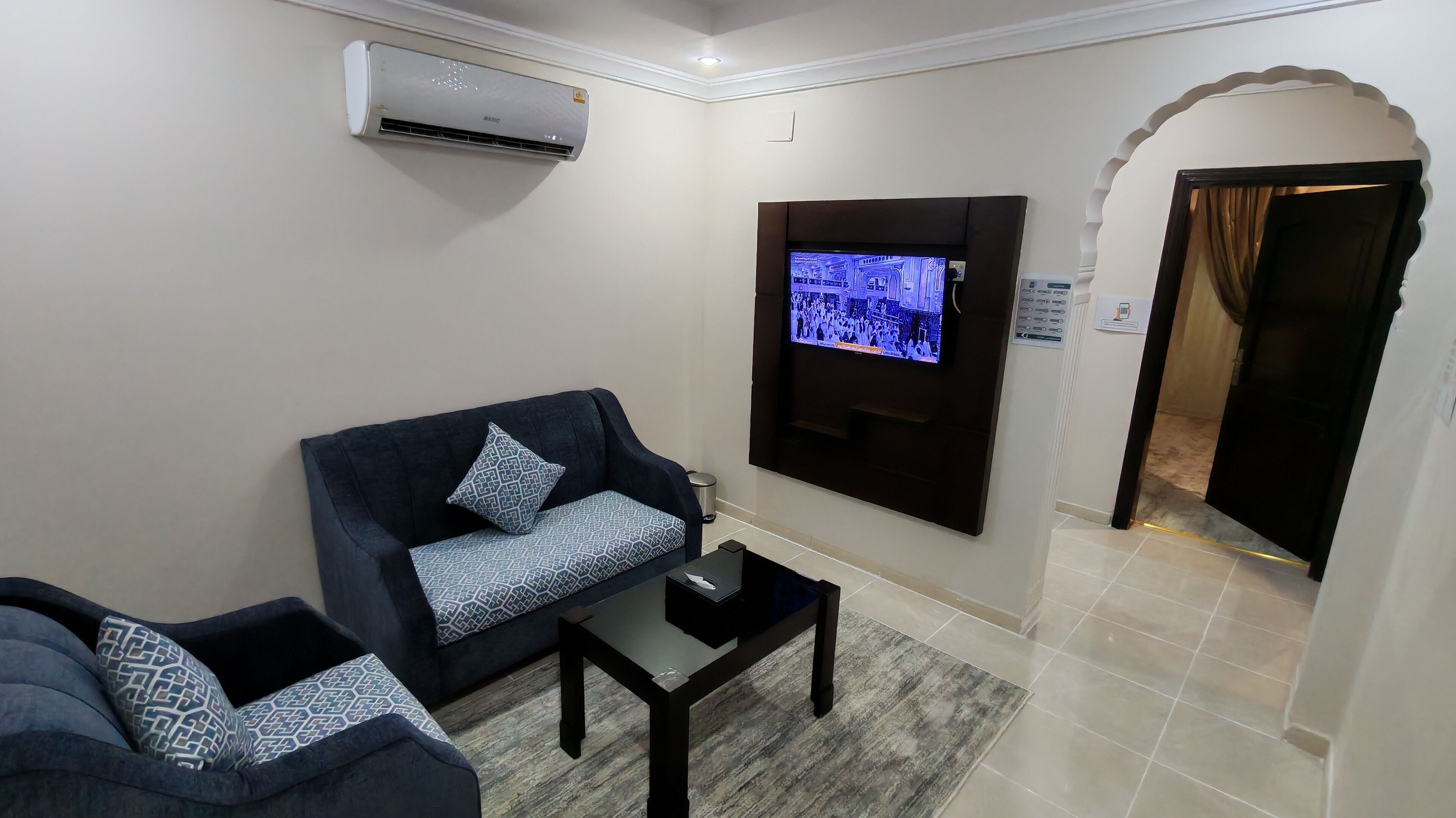 Uma sala de estar com um sofá azul escuro, uma mesinha e uma televisão na parede.