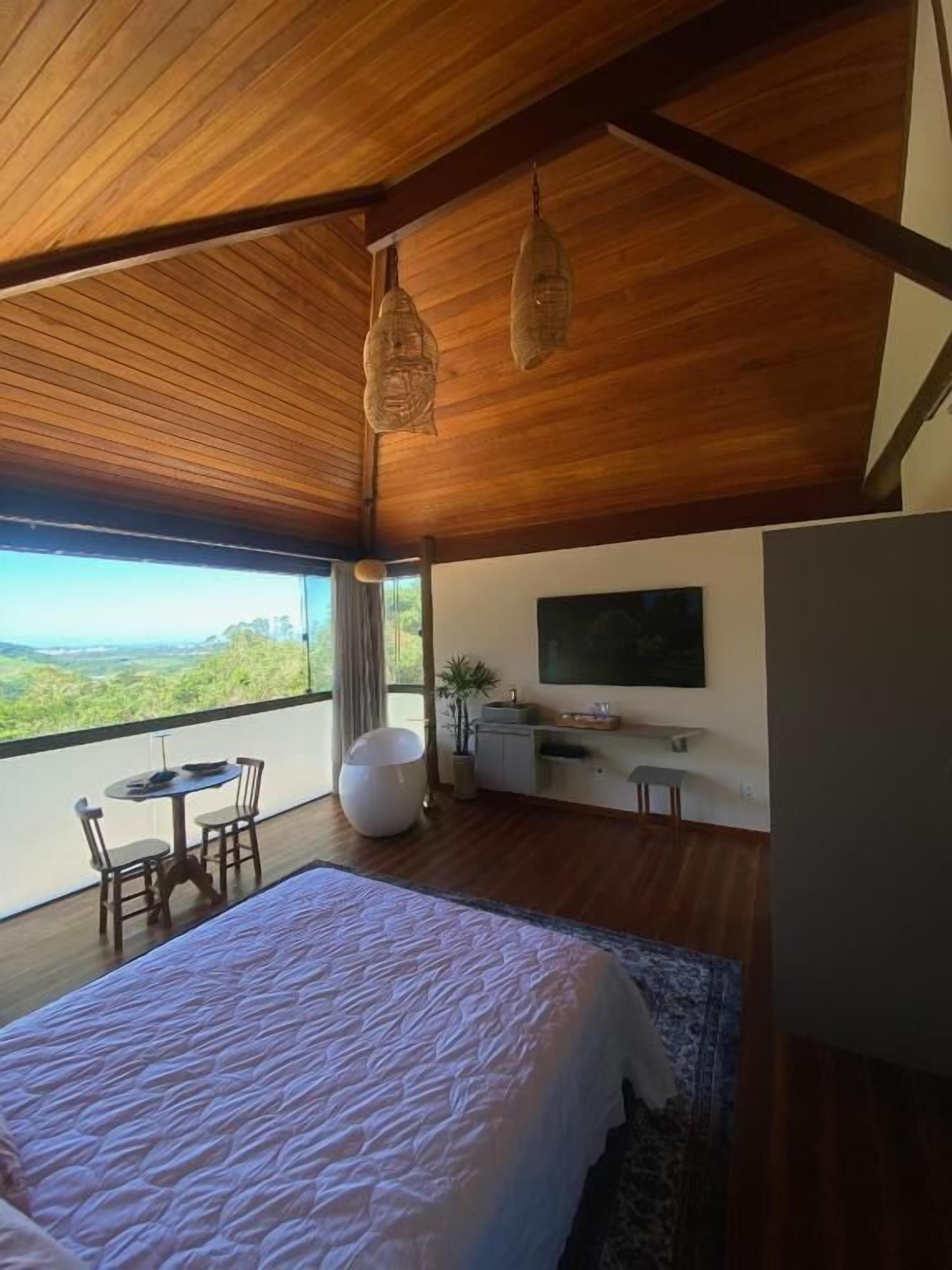 Quarto com cama grande, televisão na parede, área de refeições com mesa e cadeiras, e vista para o exterior.