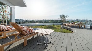 Terraço no telhado com espreguiçadeiras de madeira, uma pequena mesa e vista para a paisagem urbana.