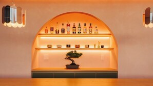Uma mesa de madeira com um bonsai como peça central, prateleiras com garrafas e uma iluminação ambiente acolhedora.