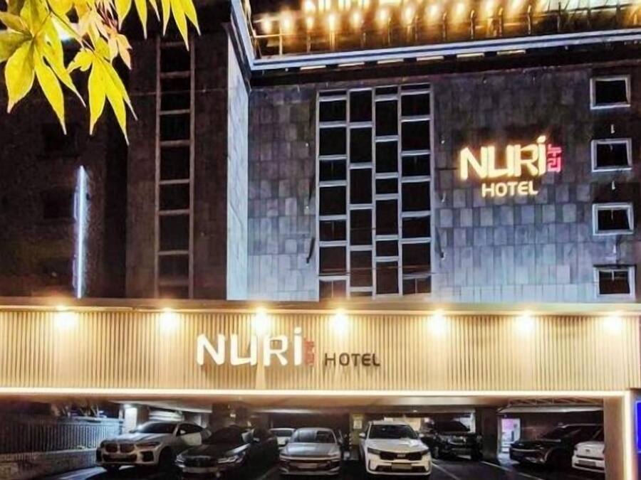 Exterior do HOTEL NURI com fachada moderna, vários espaços de estacionamento e um letreiro iluminado em destaque.