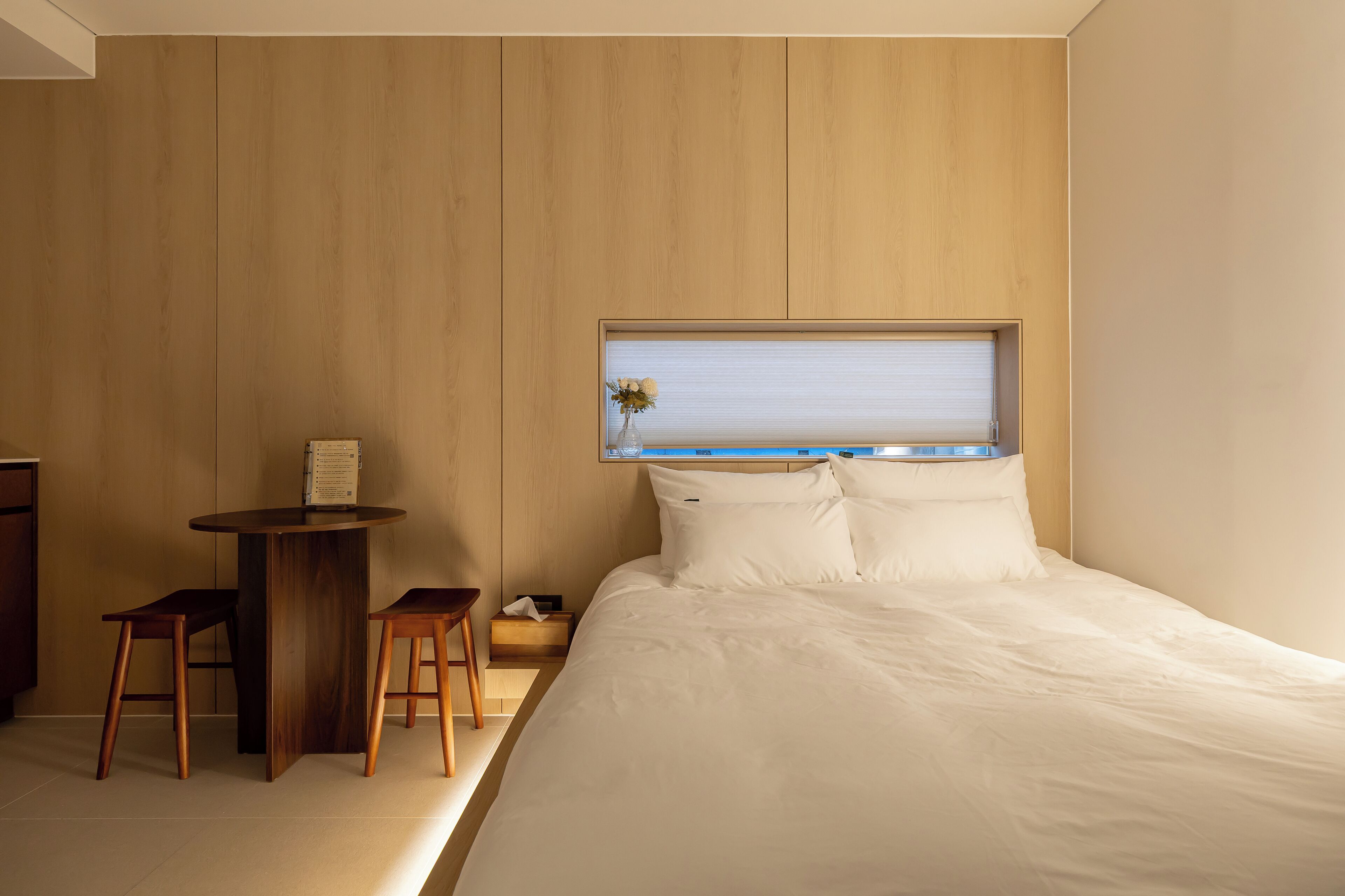 Quarto com uma cama, uma escrivaninha de madeira, dois banquinhos e uma janela com vista para o mar.