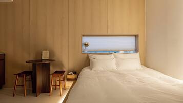 Quarto com uma cama, uma escrivaninha de madeira, dois banquinhos e uma janela com vista para o mar.
