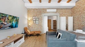 Uma sala de estar com televisão, poltrona de couro, sofá cinza, piso de madeira, parede de tijolos e um vigamento de madeira no teto.