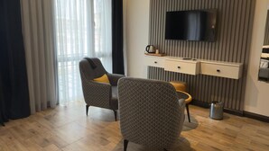 Uma sala de estar moderna com piso de madeira, duas poltronas estampadas, uma mesinha e uma televisão de tela plana fixada na parede, além de uma luminária no teto.