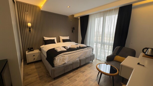 Quarto de hotel moderno com uma cama grande, uma mesa de cabeceira, uma cadeira, uma mesinha e uma janela com cortinas.