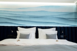 Cama com cabeceira escura, roupa de cama branca e almofadas decorativas.