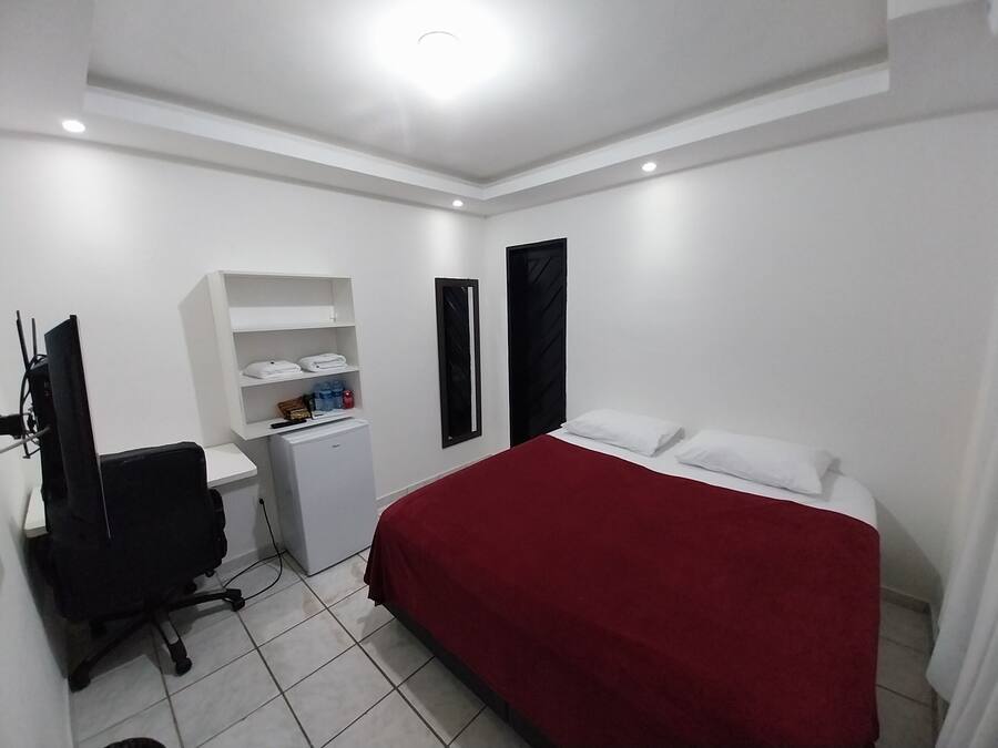 Quarto pequeno e limpo com cama, escrivaninha, mini-geladeira e prateleira com produtos de higiene pessoal.
