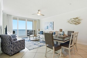 Uma sala de estar espaçosa com uma grande janela com vista para o oceano, uma mesa de jantar com cadeiras e um ventilador de teto.