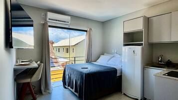 Quarto de hotel compacto com cama, pequeno espaço de cozinha, escrivaninha com cadeira, televisão fixada na parede e uma janela ampla com vista para edifícios.