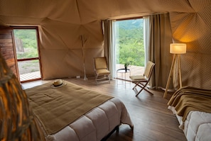 Quarto tipo tenda com cama, cadeiras, luminária e vista para o verde.