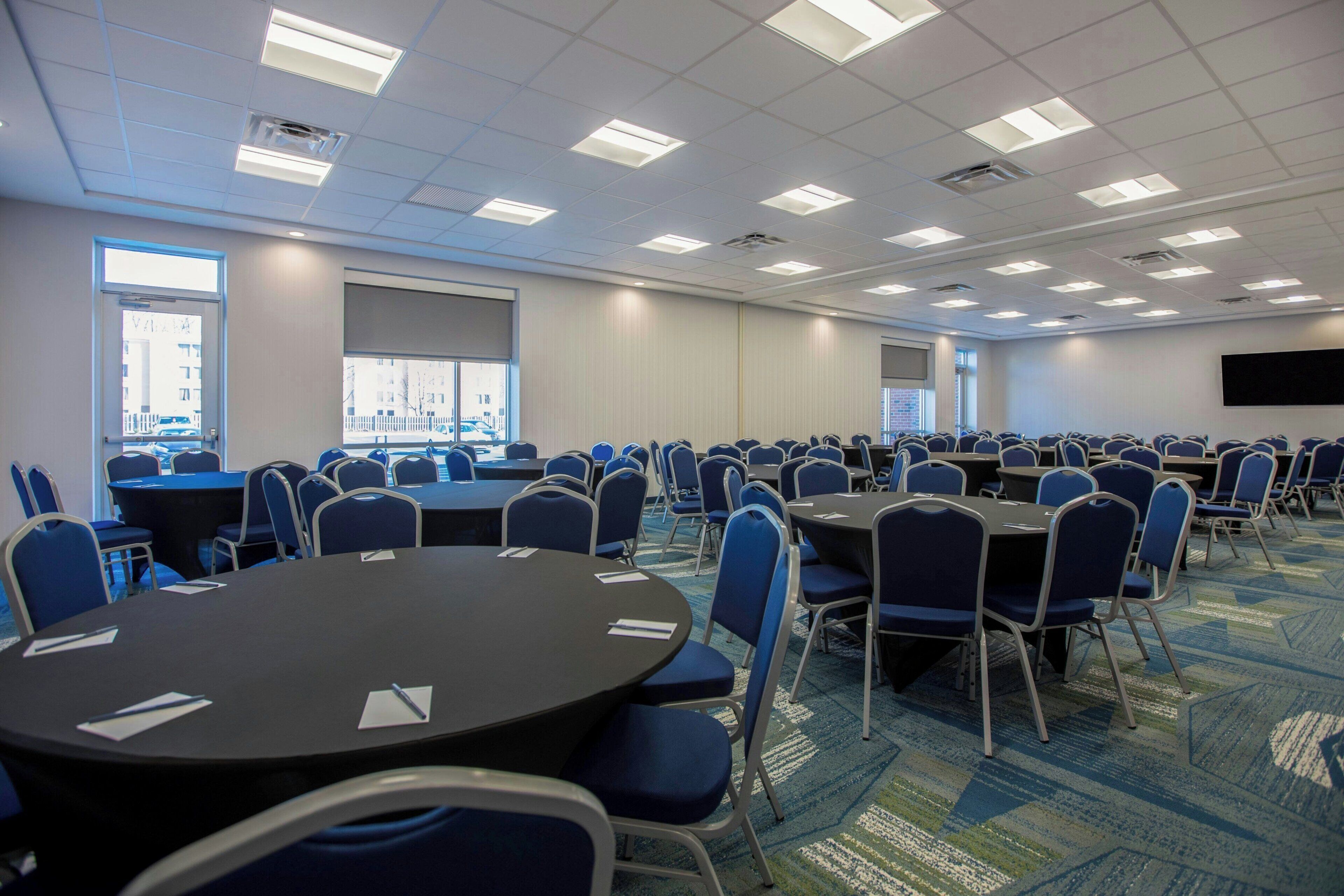 Sala de conferências com várias mesas e cadeiras, janelas amplas e piso coberto de carpete.