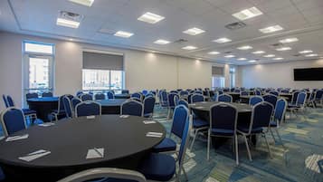 Sala de conferências com várias mesas e cadeiras, janelas amplas e piso coberto de carpete.