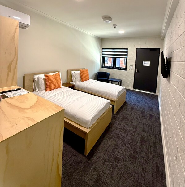 Quarto de hotel com duas camas, uma escrivaninha, uma cadeira, uma mesinha, uma janela e uma porta.