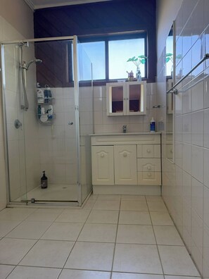 Casa de banho com chuveiro envolto em vidro, toucador branco com armários e janela sobre o espelho.