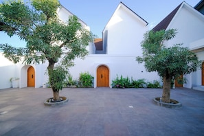 Um pátio com duas árvores, uma porta de madeira e um edifício branco com telhado inclinado.