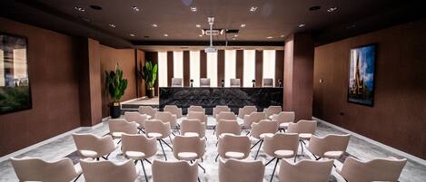 Sala de conferência com fileiras de cadeiras voltadas para uma mesa de recepção, janelas amplas e teto com iluminação embutida.