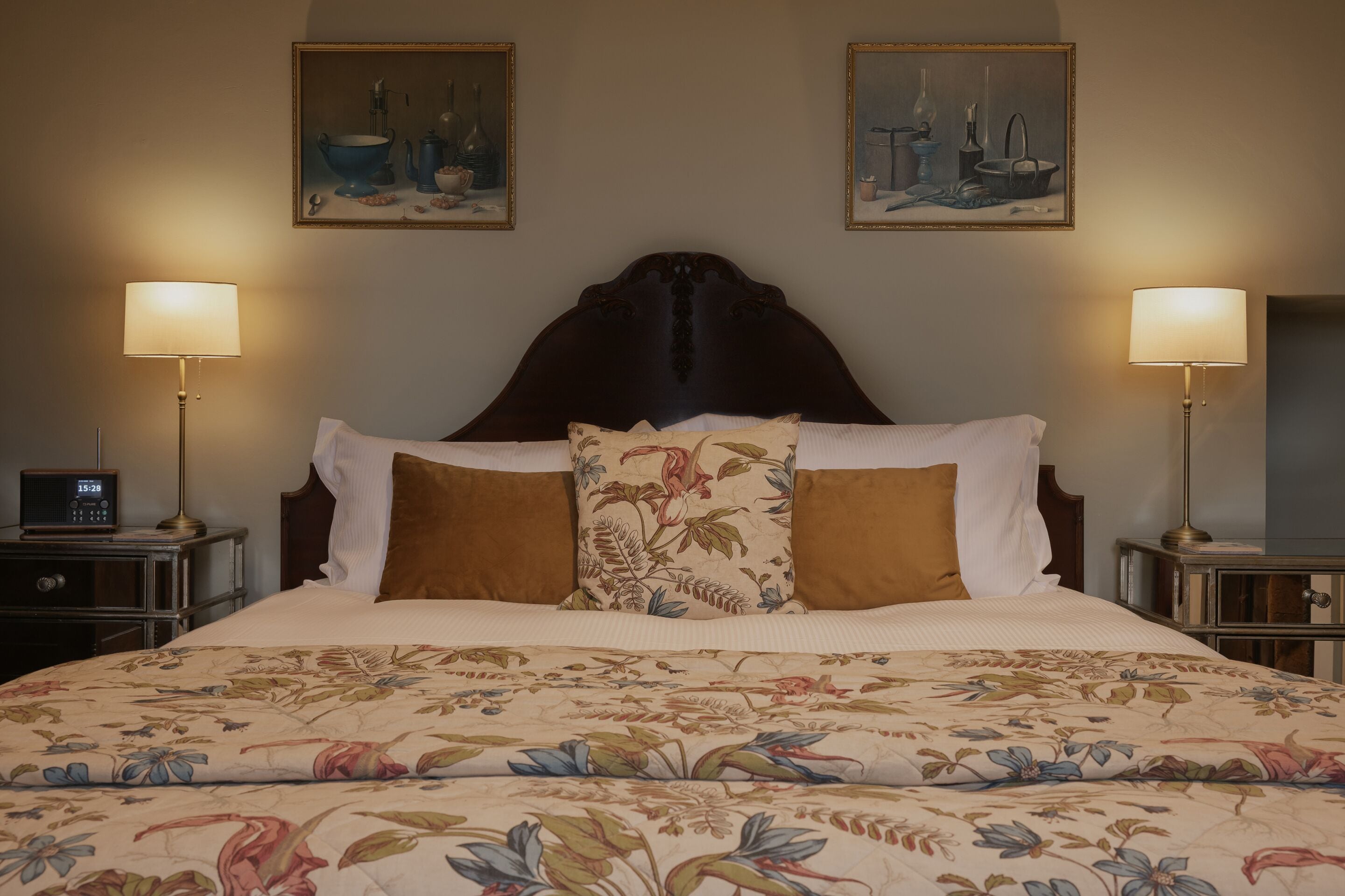Cama bem arrumada com roupa de cama florida, duas luminárias de cabeceira e dois quadros na parede.