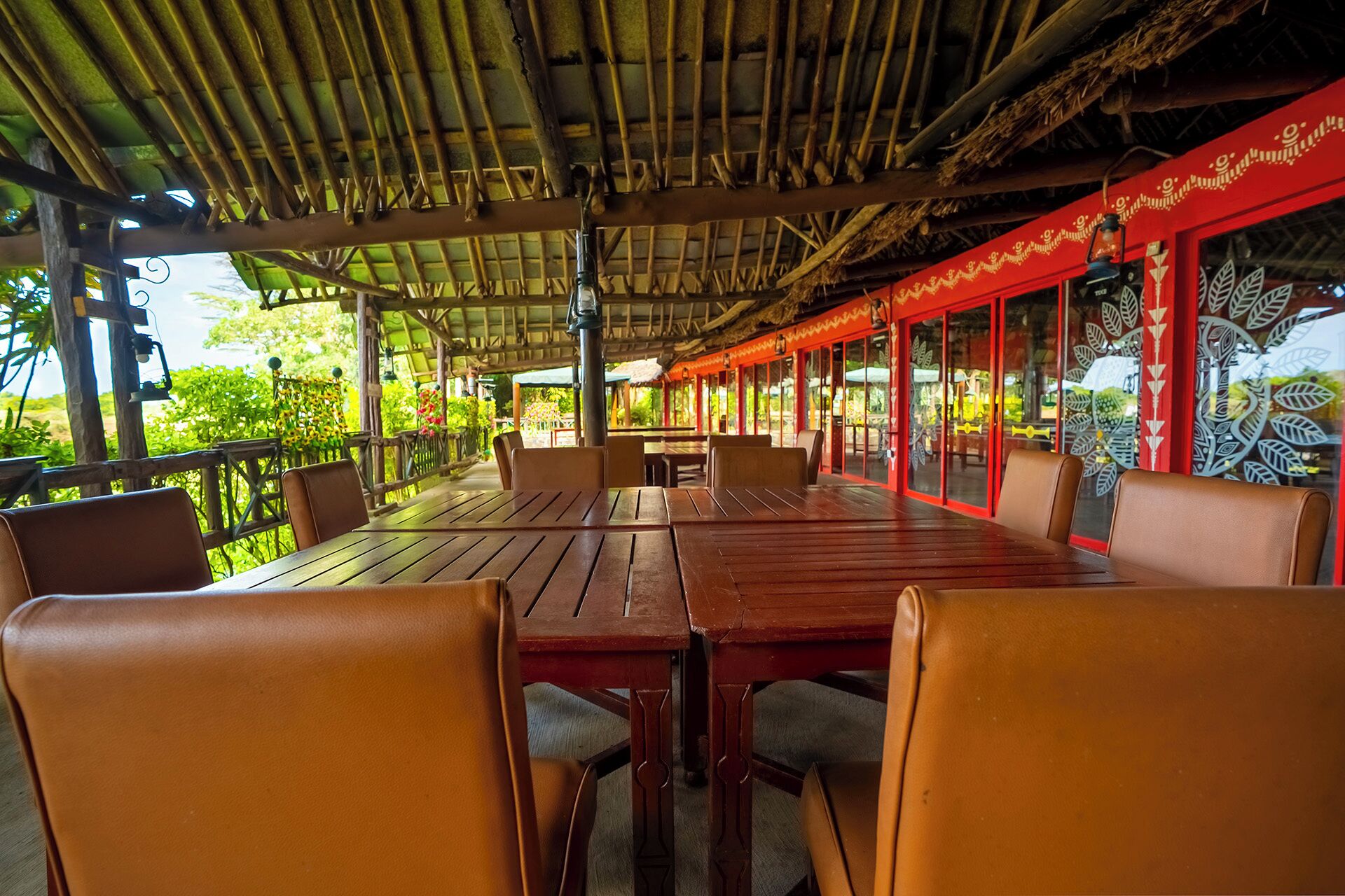 Restaurante ao ar livre com mesas e cadeiras de madeira, telhado de bambu e portas de vidro com moldura vermelha.