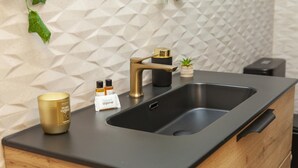 Uma cozinha com bancada preta, torneira dourada e pia preta. Há duas garrafas na bancada, uma delas com a etiqueta "Dream algarve" e outra com tampa dourada. Uma pequena planta em vaso está na bancada.