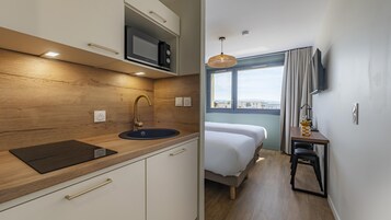 Quarto de hotel moderno com bancada de madeira, pia e micro-ondas. Inclui cama, mesa pequena e TV fixada na parede.