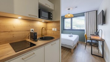 Quarto de hotel moderno com bancada de madeira, pia, micro-ondas, cama, escrivaninha e uma grande janela com vista para as montanhas.