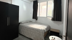 Quarto com cama, guarda-roupa, mesa pequena com objeto decorativo e janela com cortinas.
