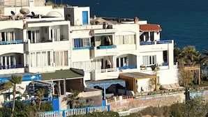 Uma área residencial costeira com edifícios brancos modernos, vista clara para o mar e paisagem montanhosa ao fundo.