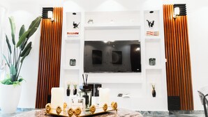 Uma sala de estar moderna com uma grande TV de tela plana na parede, uma mesa de centro redonda de mármore com objetos decorativos e uma planta em vaso.