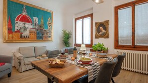 Uma área de refeições com mesa de madeira posta para uma refeição, uma pintura de paisagem urbana, um sofá e uma janela com cortinas.