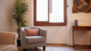 Um quarto com poltrona cinza, planta em vaso, soalho de madeira, janela com persianas, retrato emoldurado e uma pequena mesa de madeira com livros.